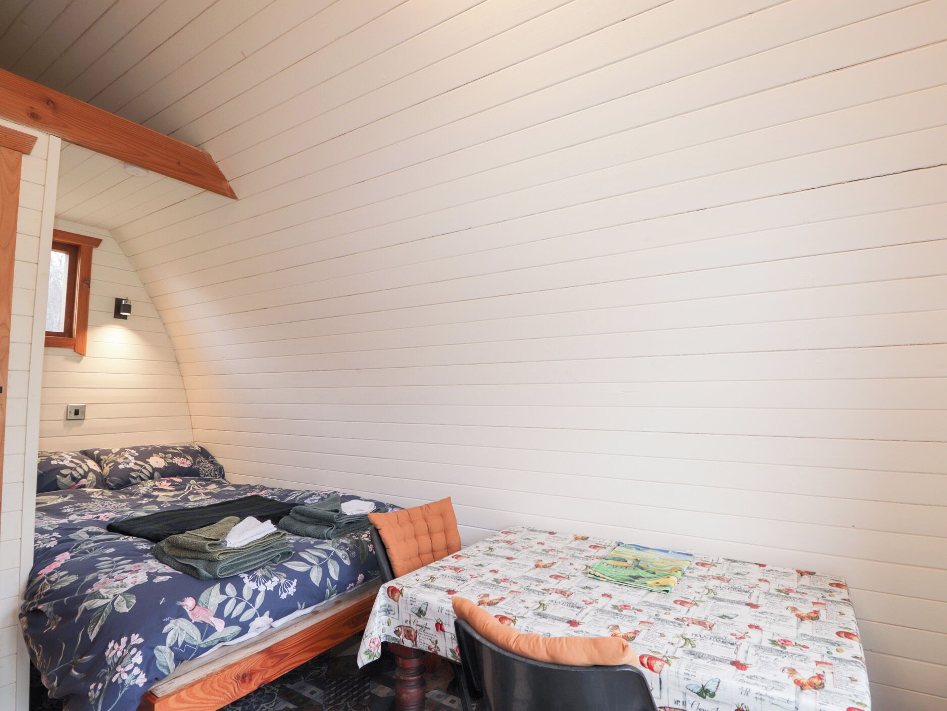 Um quarto pequeno e acolhedor, com teto inclinado, uma cama com roupa de cama florida, uma mesinha com toalha de mesa estampada com frutas e uma janela com moldura de madeira.