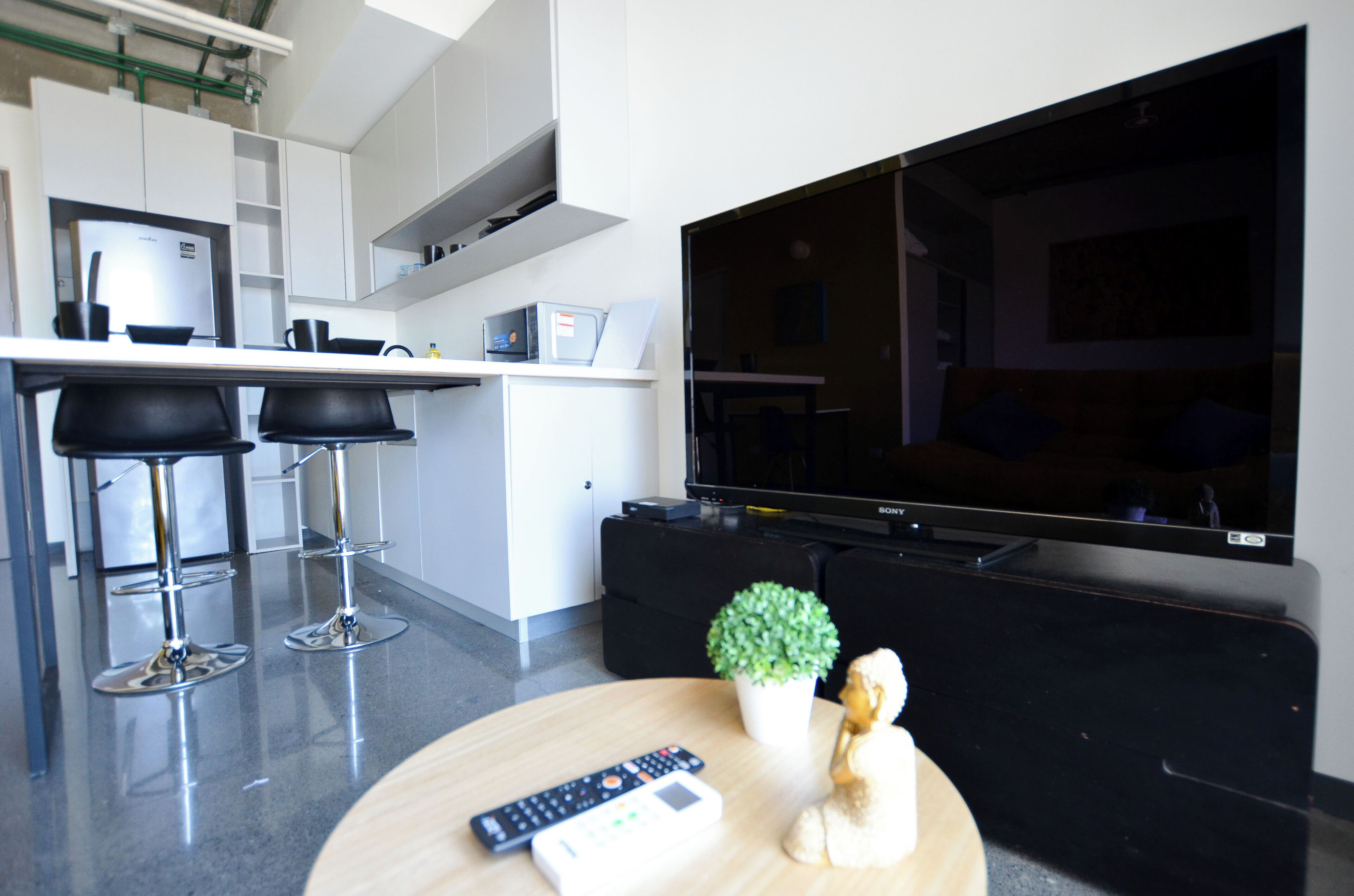 Uma sala de estar moderna com uma grande televisão de tela plana na parede, uma mesa de centro com um controle remoto, uma planta decorativa e uma pequena estatueta.