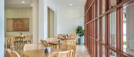 Uma sala de jantar de hotel com mesas e cadeiras de madeira, uma grande planta em vaso e uma placa fixada na parede.