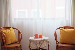 Duas cadeiras de vime com almofadas amarelas, uma pequena mesa redonda com toalha de renda e duas chávenas laranjas sobre a mesa.