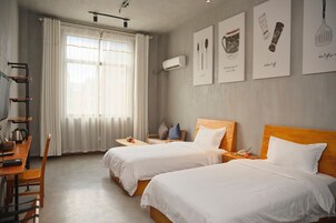 Quarto de hotel com duas camas, uma escrivaninha com cadeira, uma mesinha e uma janela com cortinas. Há obras de arte nas paredes e um ar condicionado.
