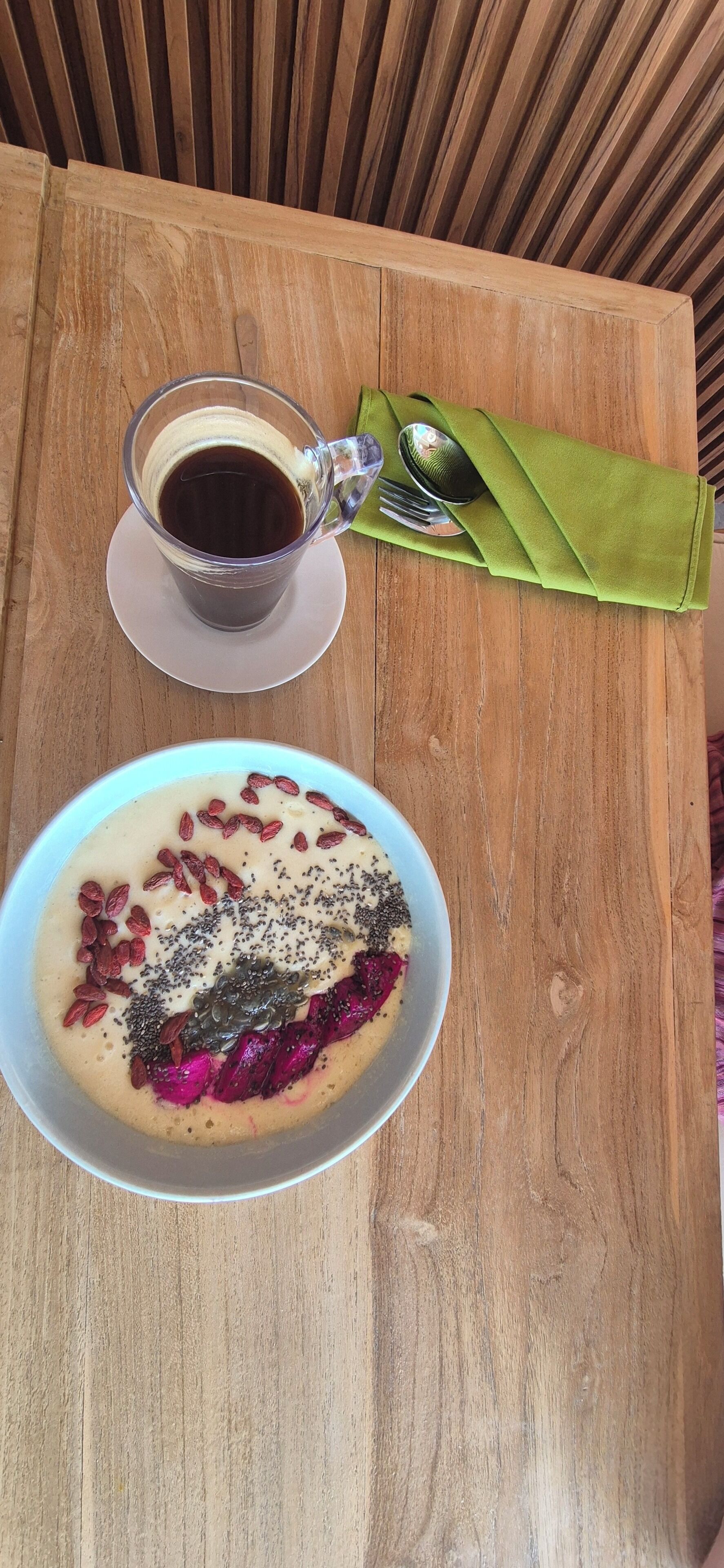 Uma mesa de madeira com uma chávena de vidro transparente com café e uma tigela de comida com sementes e fruta.