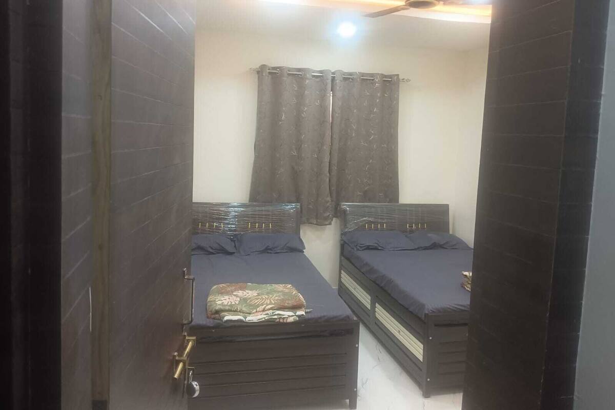 Um quarto pequeno, com pouca luz, com duas camas de solteiro, um ventilador de teto e uma janela com cortinas.