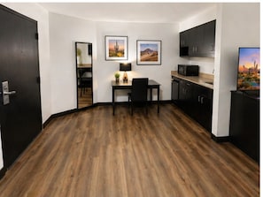 Quarto compacto com piso de madeira, mesa de jantar com cadeiras, pequena luminária, quadros nas paredes, micro-ondas e armários.