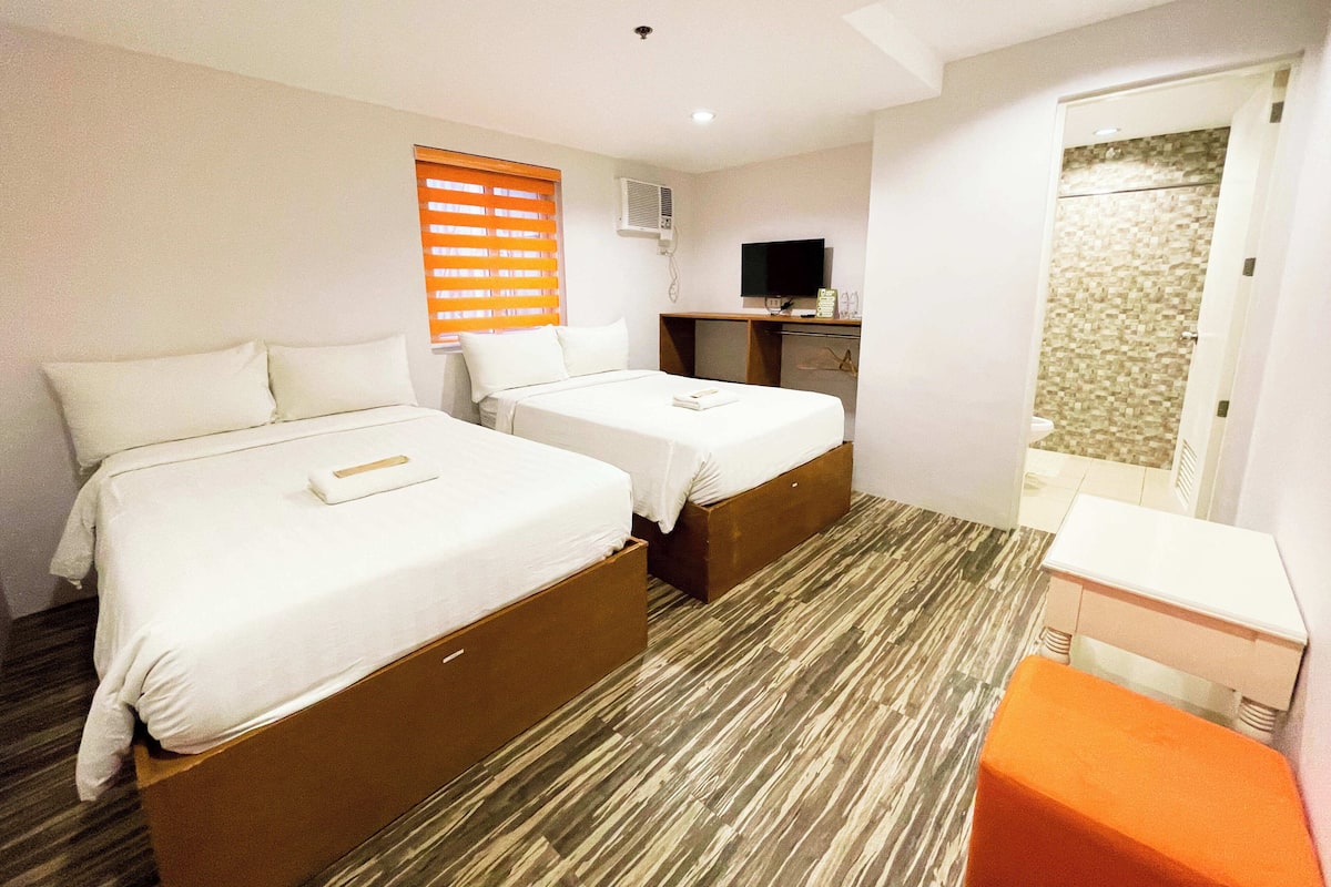 Quarto de hotel com duas camas, uma escrivaninha, televisão e zona de chuveiro.