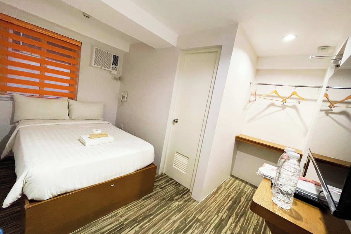 Quarto de hotel com cama, escrivaninha, laptop, garrafa de água e cabideiro com cabides.