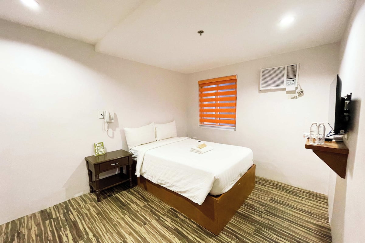 Quarto de hotel com cama, mesa de cabeceira, telefone, janela com persianas, ar condicionado e televisão sobre uma estante.