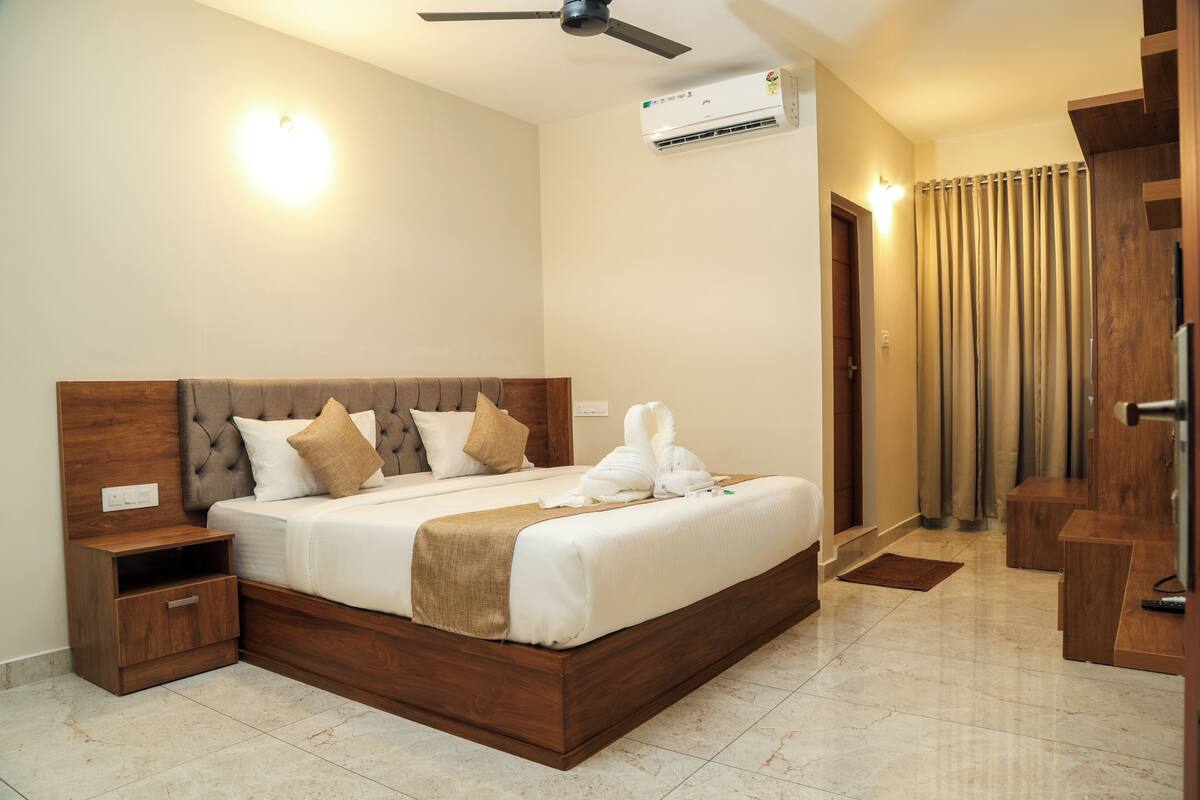 Quarto de hotel com uma cama grande, uma mesa de cabeceira, um guarda-roupa de madeira, um ventilador de teto e um aparelho de ar condicionado.