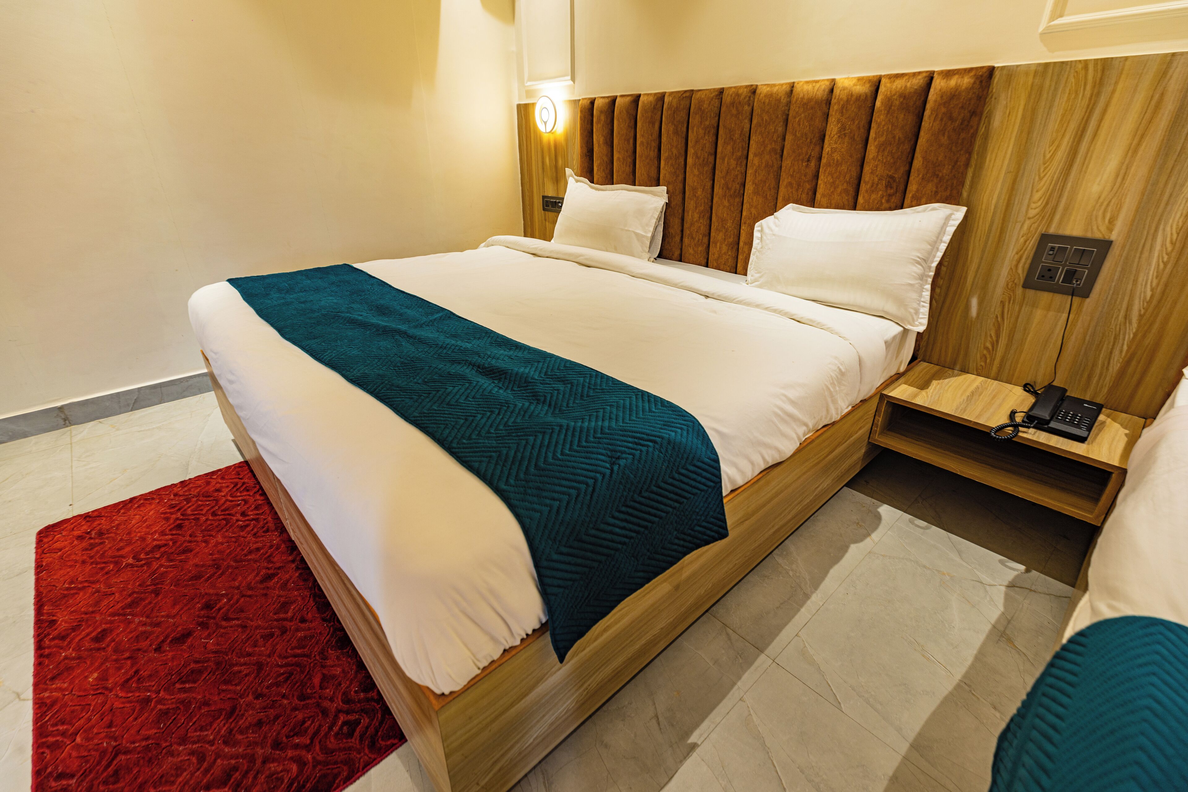 Cama bem arrumada com manta azul-turquesa, cabeceira de madeira e mesa de cabeceira com um telefone.
