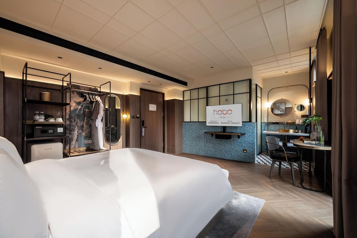 Quarto de hotel com uma cama grande, uma escrivaninha com cadeira, minibar, guarda-roupa e banheiro com espelho.