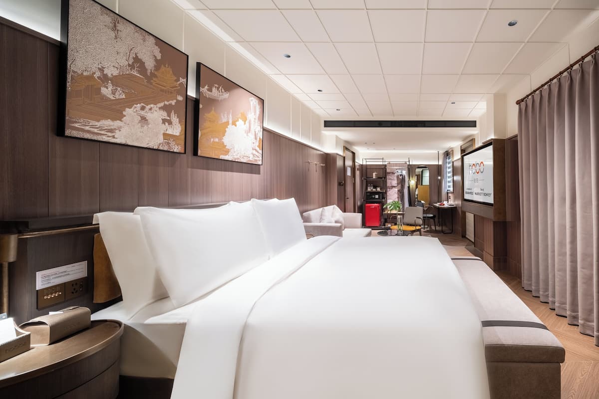 Quarto de hotel com uma cama grande, uma escrivaninha, uma cadeira, uma mesinha e um refrigerador vermelho, além de obras de arte nas paredes.