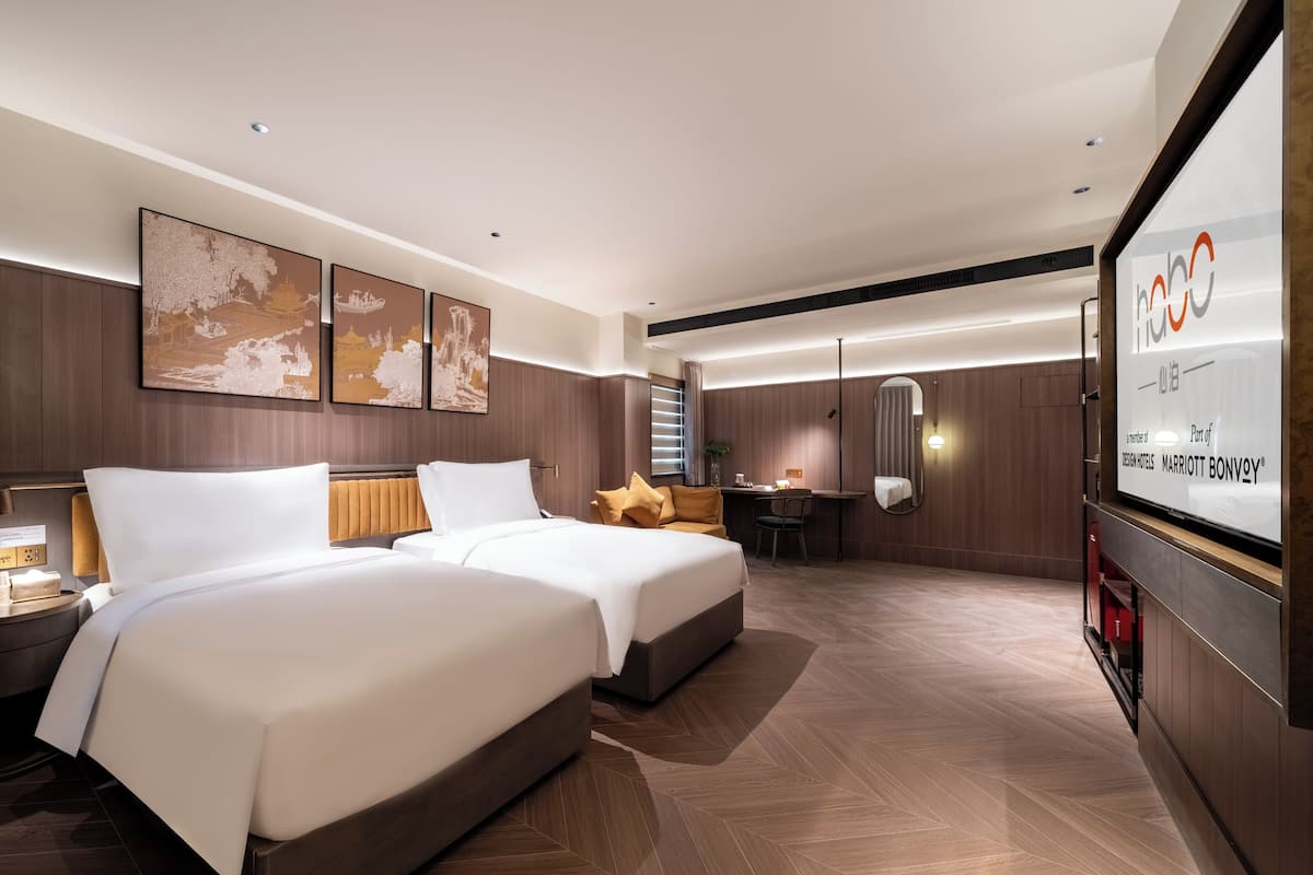 Quarto de hotel moderno com duas camas, uma escrivaninha, uma cadeira e um espelho grande.