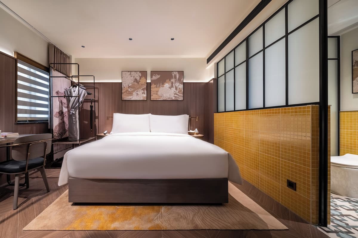 Quarto de hotel moderno com uma cama grande, uma mesa com cadeira, toucador e uma janela ampla com persianas.