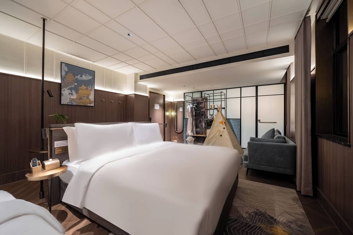 Quarto de hotel com uma cama grande, área de estar com uma poltrona, guarda-roupa, mesa de cabeceira com abajur e um quadro na parede.