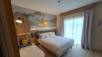 Quarto de hotel com cama, escrivaninha, cadeira, luminária e uma grande janela com cortinas.
