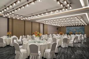 Uma sala de banquetes com mesas redondas, cadeiras brancas e iluminação pendente.