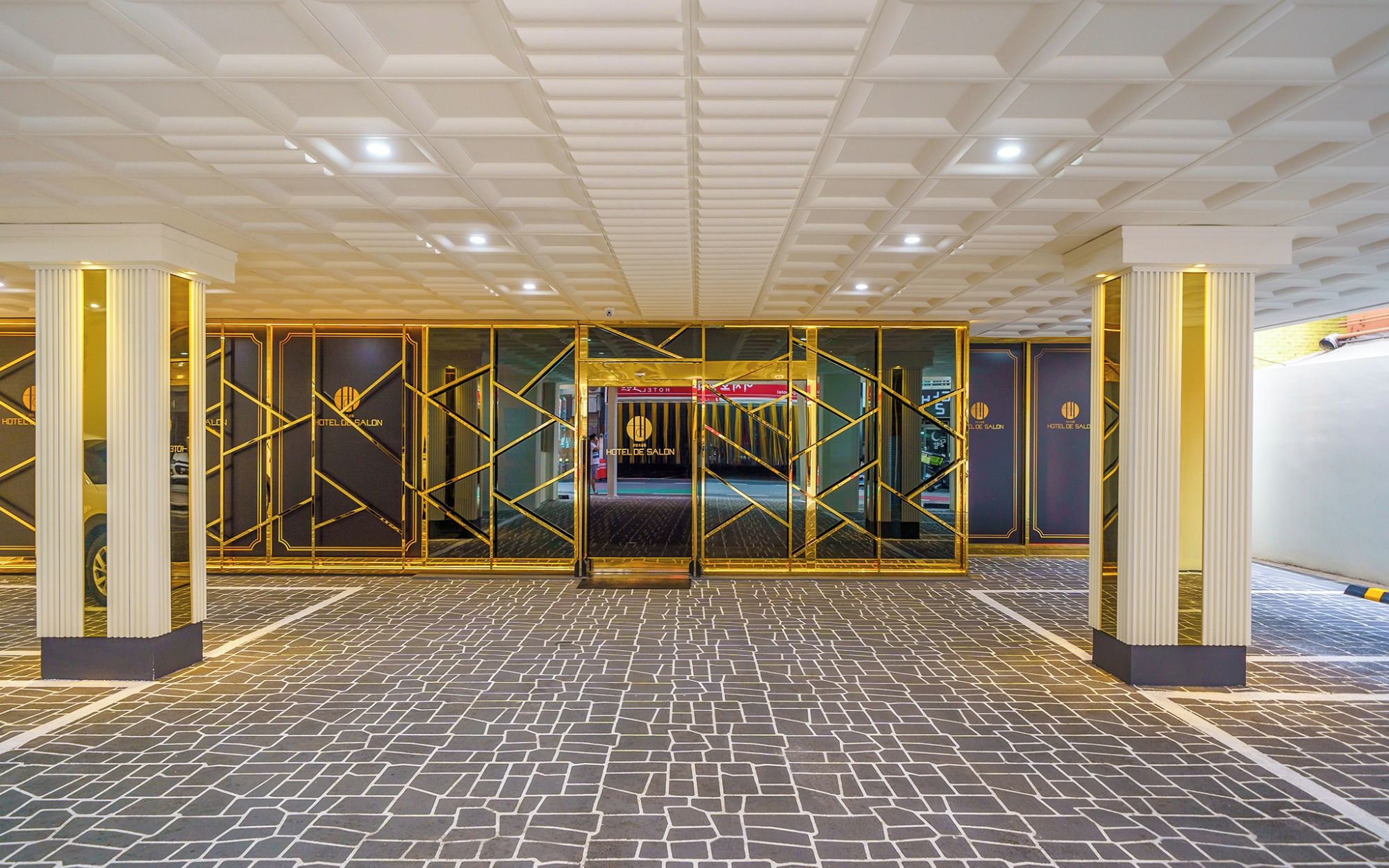 Entrada de hotel com piso de padrão geométrico, porta central emoldurada a dourado e teto com iluminação embutida.