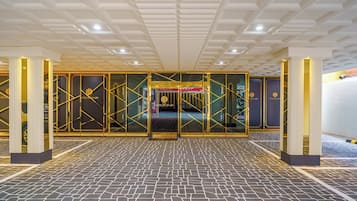 Entrada de hotel com piso de padrão geométrico, porta central emoldurada a dourado e teto com iluminação embutida.