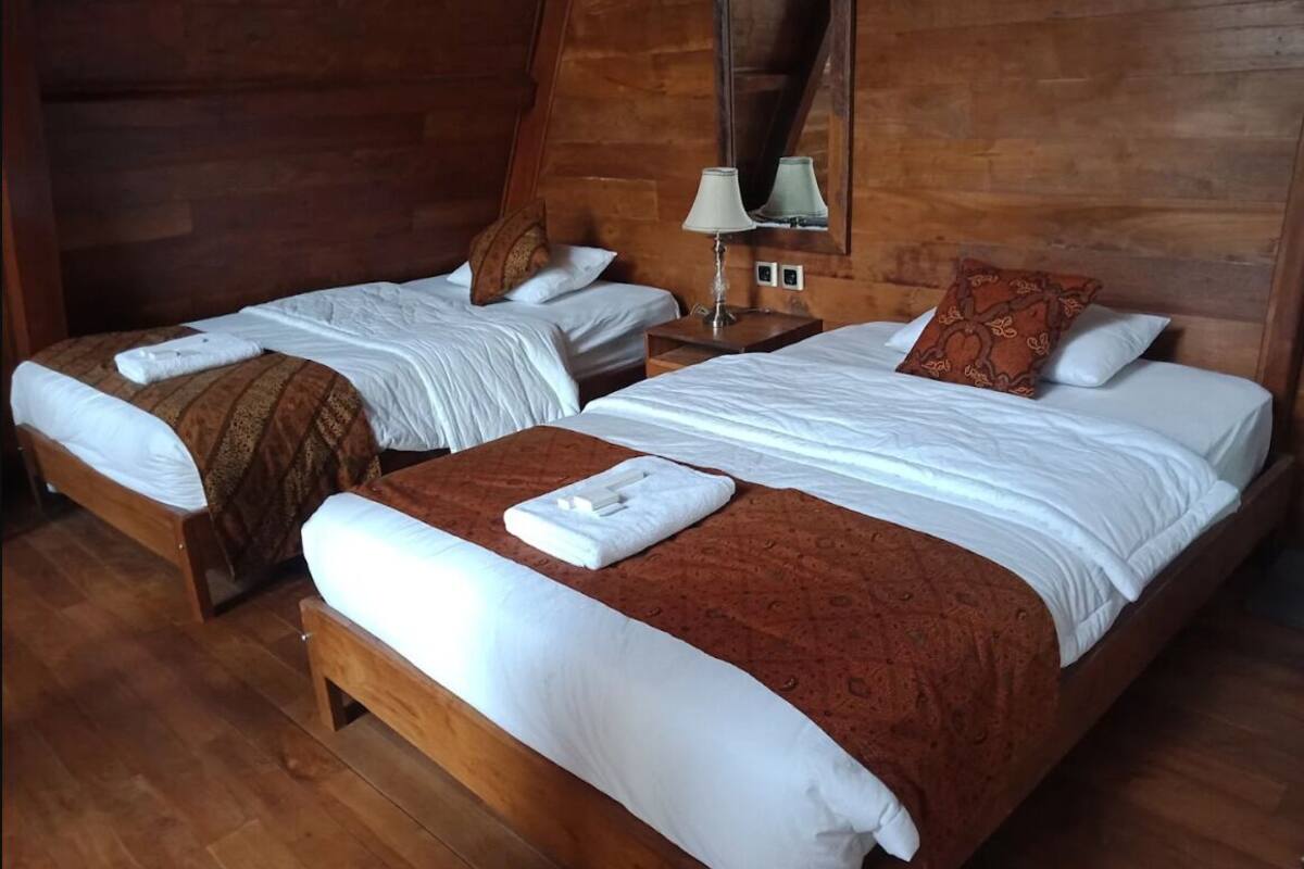 Quarto com duas camas de madeira, cada uma com lençóis brancos e cabeceira marrom. Há uma mesa de cabeceira com uma luminária entre as camas.