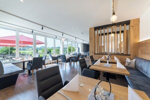 Restaurante moderno com grandes janelas, mesas de madeira e cadeiras pretas.