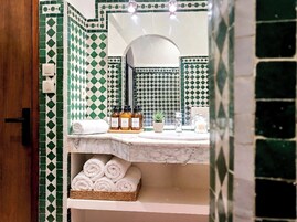 Um banheiro com painel de azulejos quadriculado em verde e branco, bancada de mármore e um espelho acima. Há toalhas, uma pequena planta e artigos de higiene pessoal na bancada.