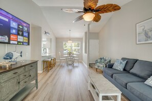 Uma sala de estar moderna com um ventilador de teto, uma TV de tela plana, um espaço de jantar com um lustre e um sofá cinza.