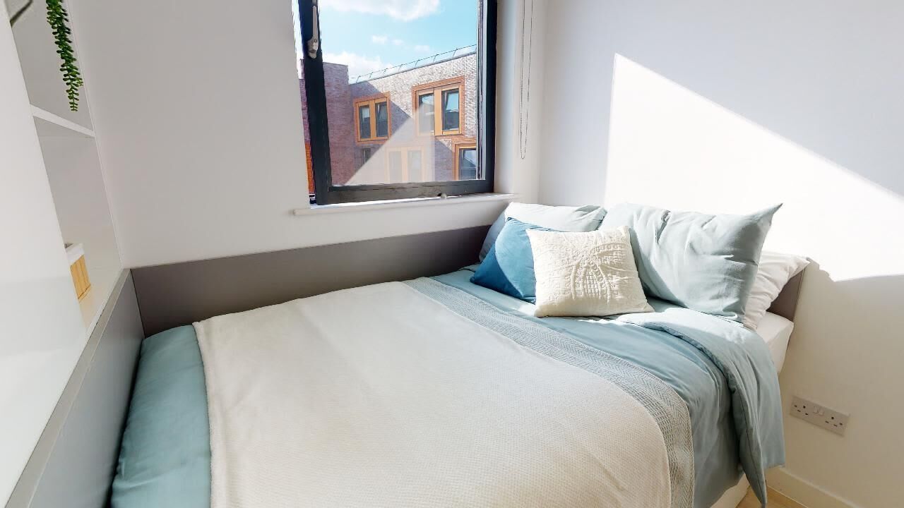 Uma cama bem arrumada com um cobreleito branco e azul, uma janela com vista para edifícios e uma pequena planta à esquerda.