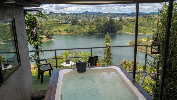 Varanda com banheira de hidromassagem com vista para um lago, cercada por vegetação e montanhas.