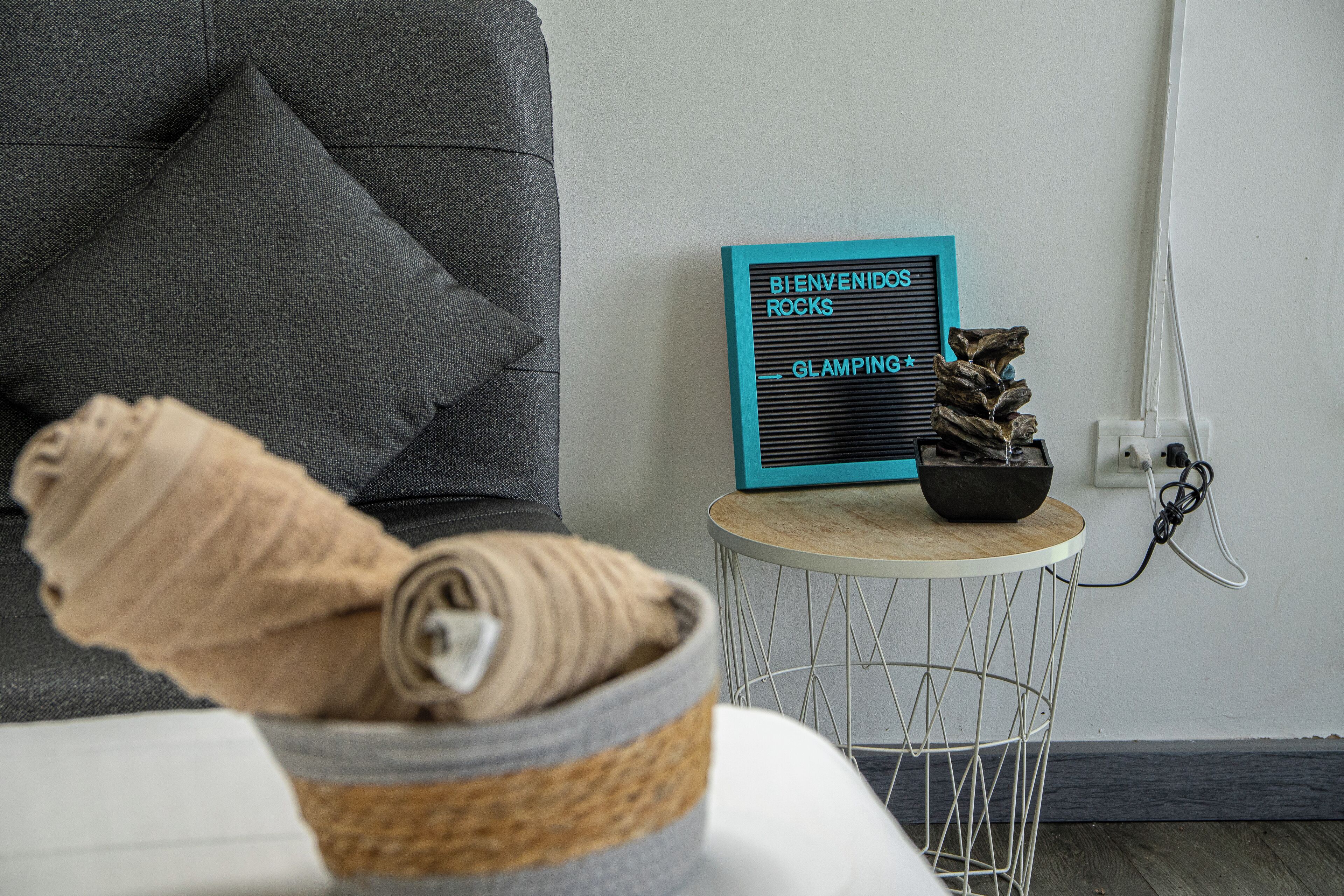 Quarto com sofá cinza, mesa redonda pequena e uma cesta de vime, além de uma placa na parede.