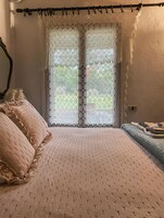 Quarto com cama, almofadas, mesa de cabeceira e janela com cortinas de renda.