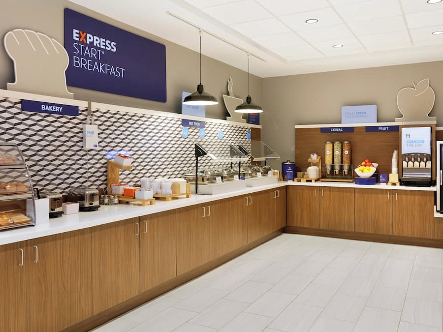 Uma área de café da manhã com uma placa indicando 'EXPRESS START BREAKFAST', uma seção de padaria e um balcão com diversos itens alimentícios e um display de menu digital.
