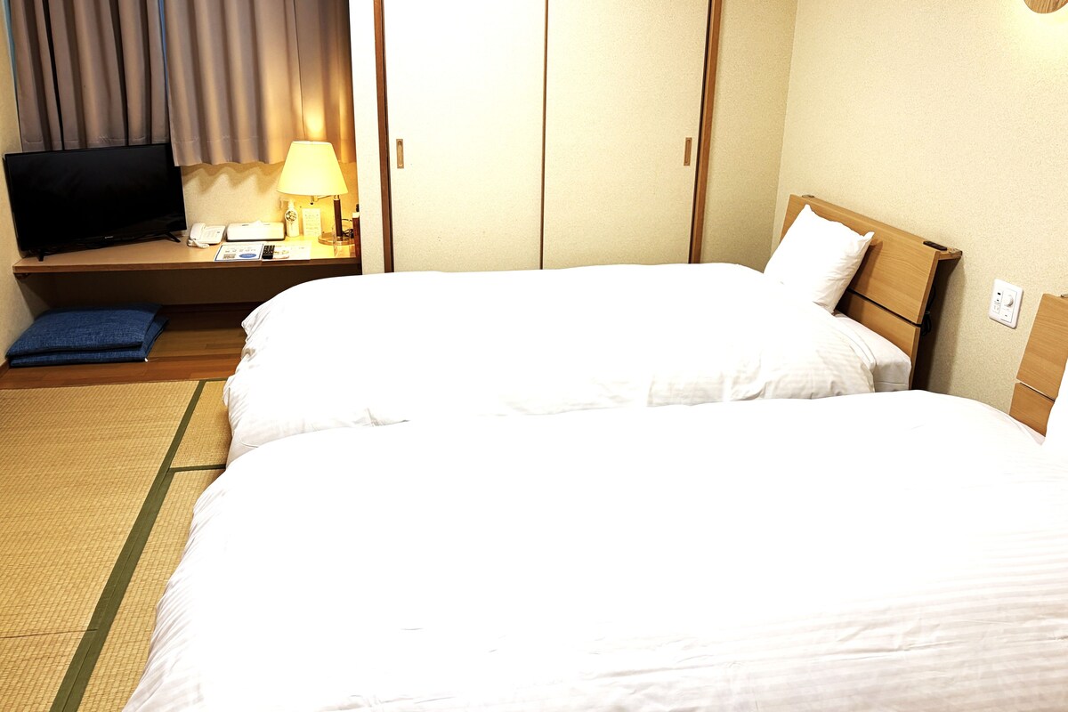 Quarto de hotel com duas camas, uma televisão, uma luminária e uma mesa de trabalho com telefone e bloco de notas.