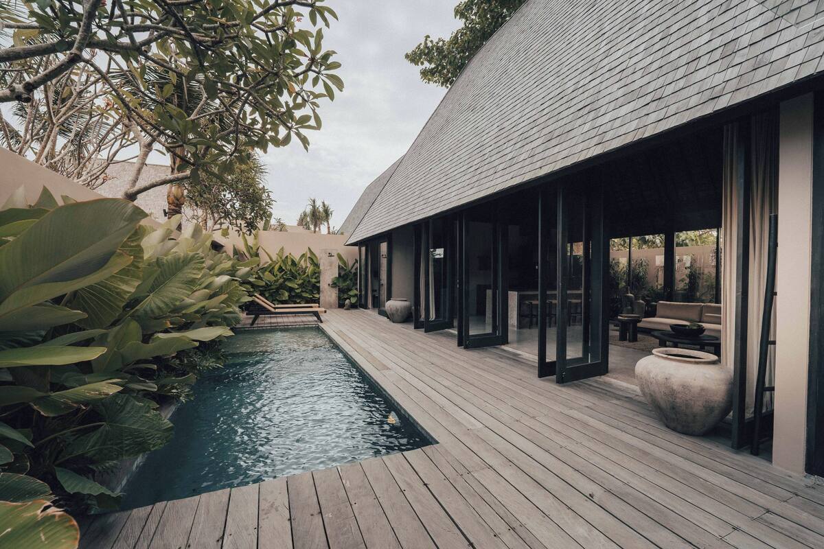 Uma área externa moderna com um deck de madeira, um pequeno lago ornamental e uma área de estar coberta.