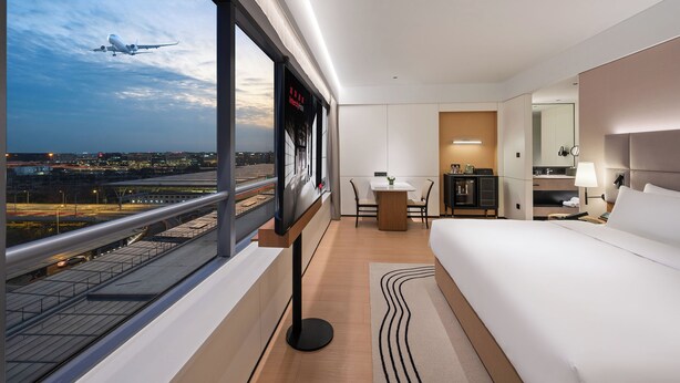 Quarto de hotel moderno com uma grande janela com vista para um aeroporto, uma cama, uma mesa de escritório e uma pequena área de refeições.