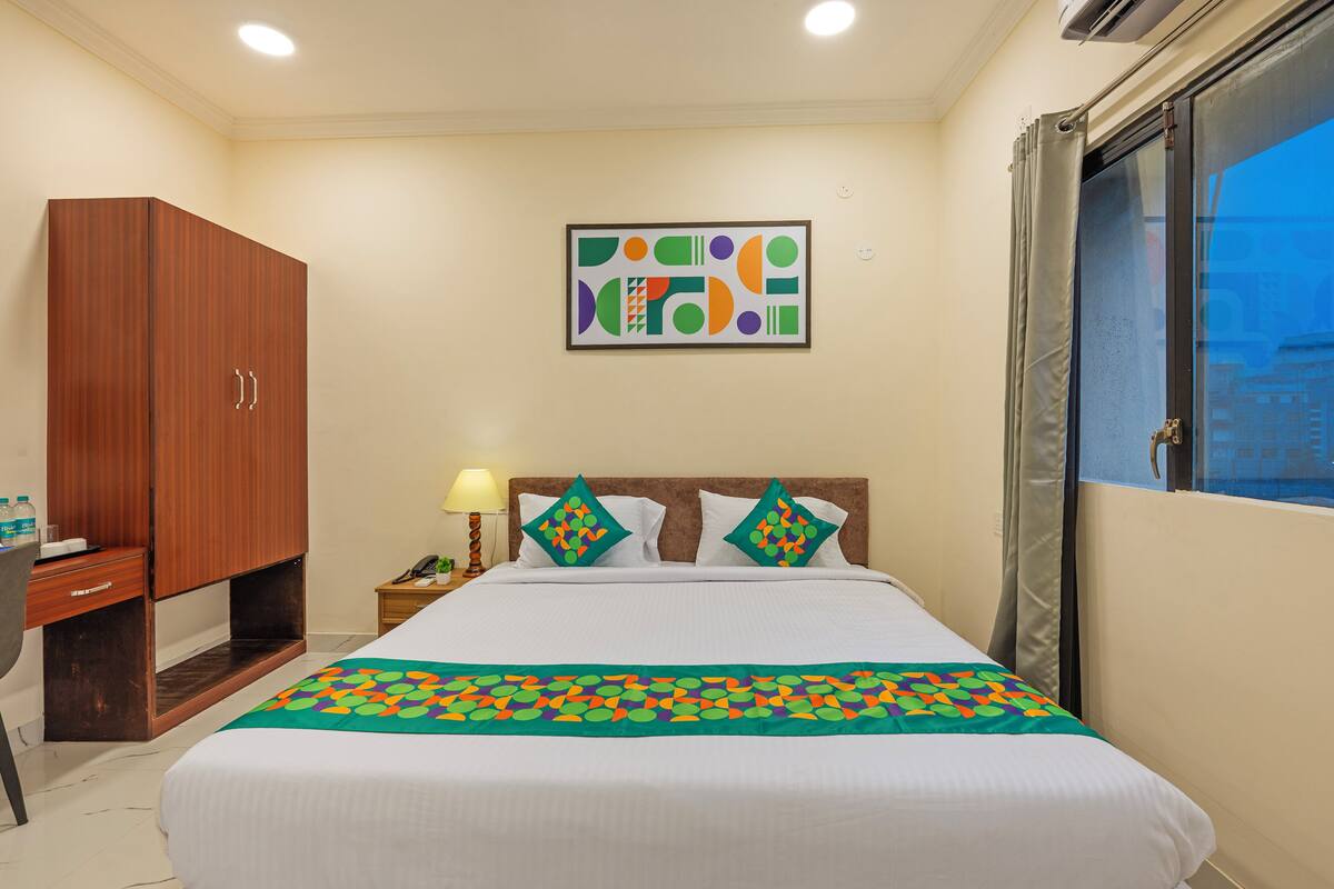 Quarto de hotel com cama, escrivaninha, guarda-roupa, mesa de cabeceira, luminária e janela com cortinas.