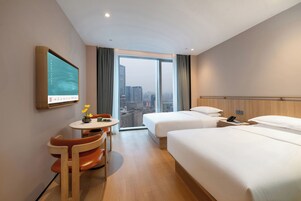 Quarto de hotel com duas camas, uma escrivaninha com cadeira, televisão fixada na parede e uma grande janela com vista para a cidade.