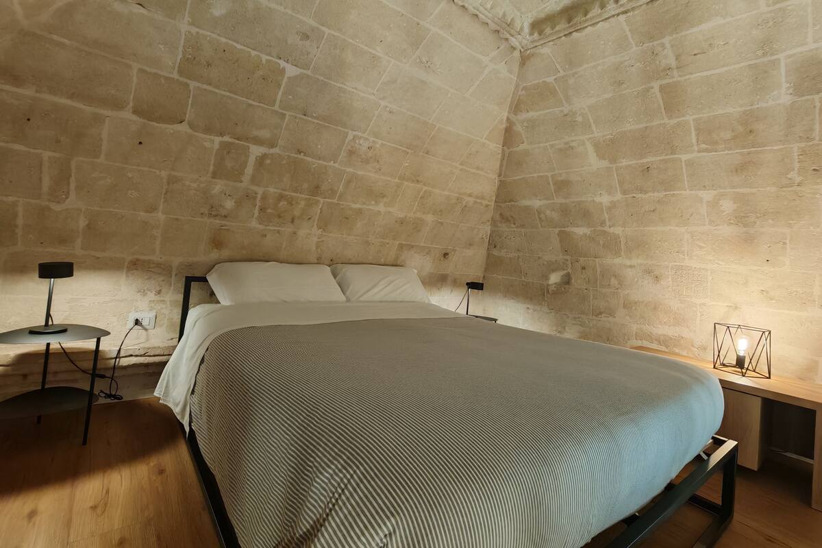 Quarto com parede de pedra, cama com manta listrada, mesa de cabeceira com abajur e escrivaninha de madeira.
