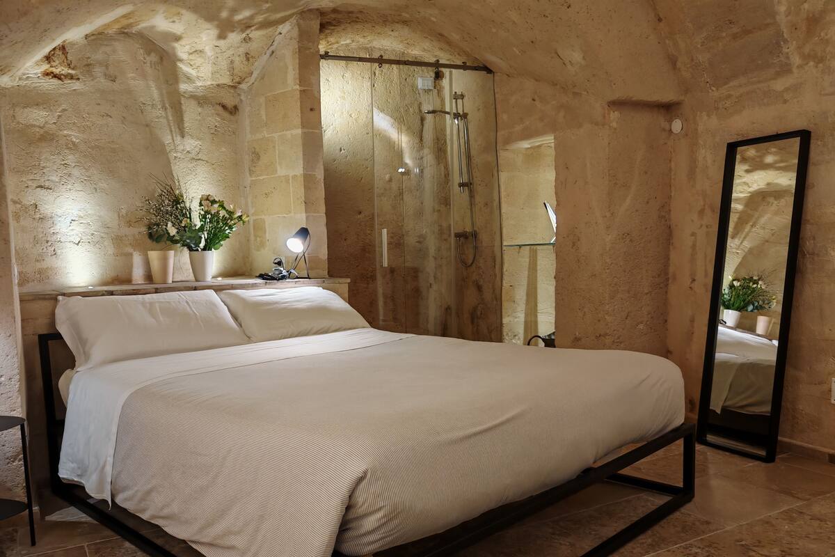 Quarto com cama grande, mesa de cabeceira, espelho e chuveiro.