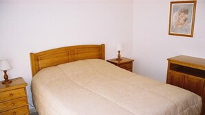 Quarto com cama de madeira, mesa de cabeceira com abajur, pequeno armário e quadro na parede.