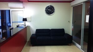 Uma sala de estar com um sofá preto, um relógio de parede, uma mesinha com uma luminária e uma porta de vidro deslizante.