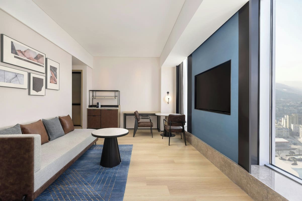 Uma sala de estar moderna com um sofá, uma mesa de jantar e cadeiras, uma TV fixada na parede e uma grande janela com vista para a cidade.