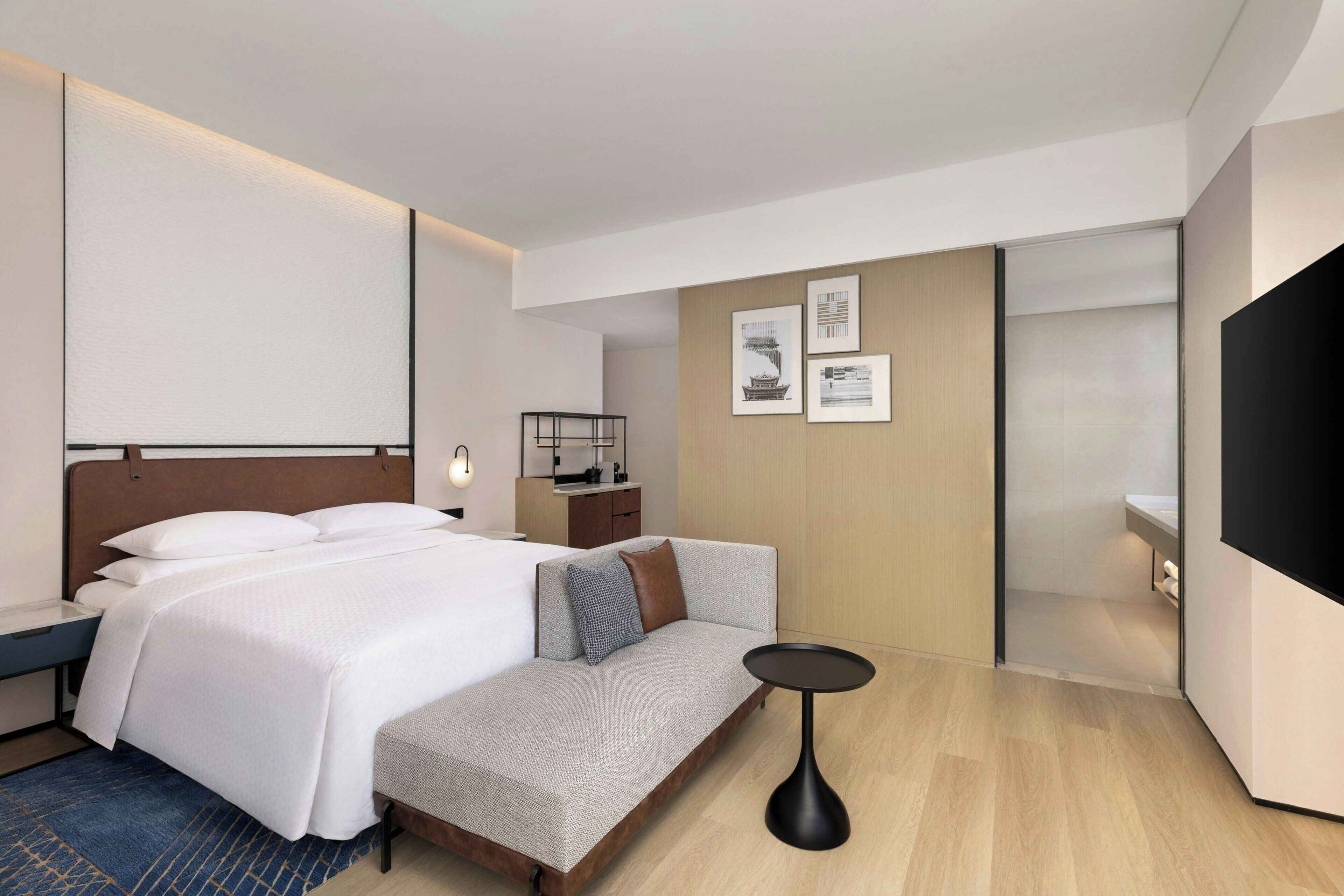 Quarto de hotel moderno com cama, sofá, mesinha e televisão.