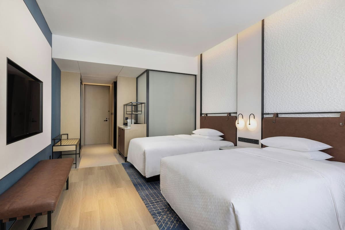 Quarto de hotel com duas camas, um banco, uma escrivaninha e uma televisão.