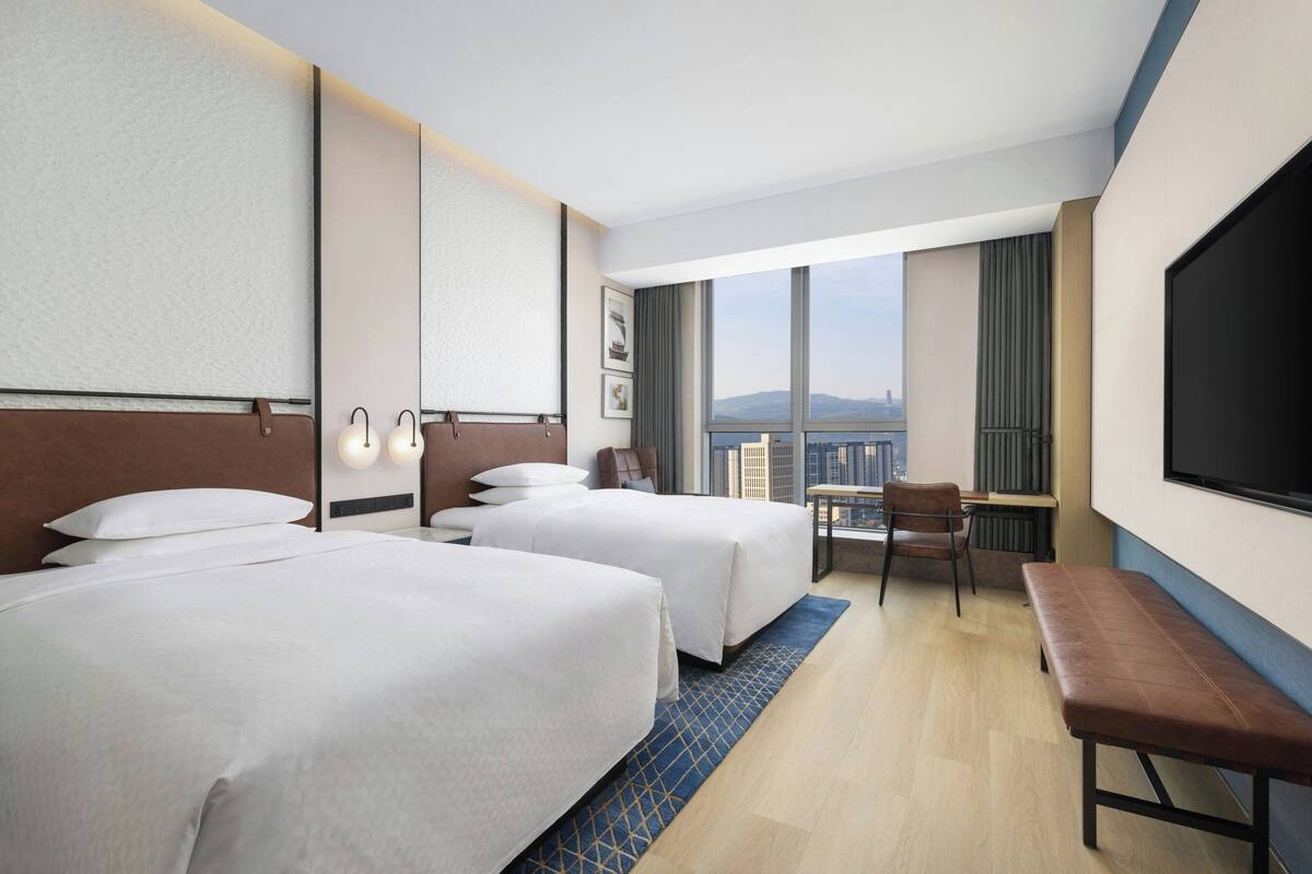 Quarto de hotel com duas camas, uma escrivaninha, uma cadeira, televisão e vista para a cidade.