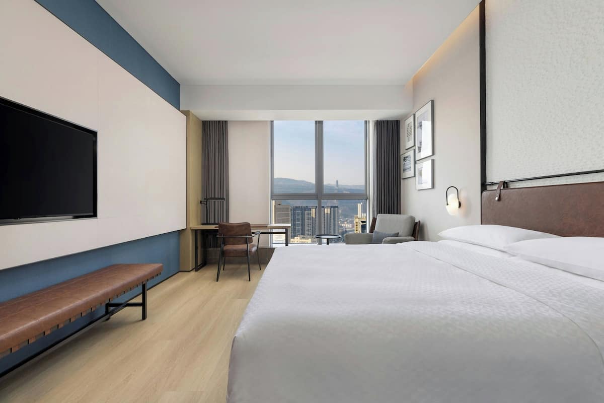 Quarto de hotel com uma cama grande, um banco, uma escrivaninha, uma cadeira, uma televisão e vista para a cidade por grandes janelas.