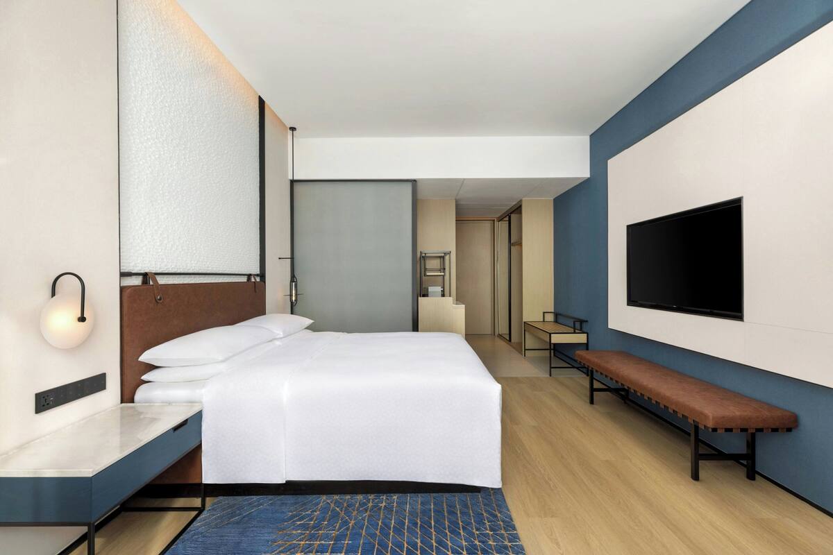 Quarto de hotel moderno com cama, mesa de cabeceira, banco, TV e vista para o corredor.