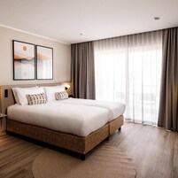 Cama de casal com cabeceira, duas mesas de cabeceira, tapete redondo de fibra natural e uma janela grande com cortinas.