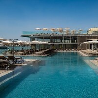 Uma área de piscina com espreguiçadeiras e guarda-sóis, uma piscina azul cristalina e um edifício moderno com grandes janelas.