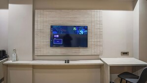 Quarto moderno com TV de tela plana fixada em parede de painel de madeira, uma escrivaninha com cadeira e uma pequena prateleira com duas garrafas.