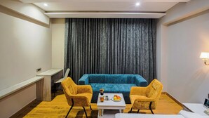 Quarto com sofá azul, duas poltronas amarelas, uma mesinha com uma travessa de fruta e uma bebida, e um abajur na parede.